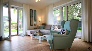 Uma sala de estar espaçosa com uma poltrona verde-clarinha, um sofá bege com almofadas estampadas, uma mesa de centro de vidro e janelas amplas com vista para o exterior.