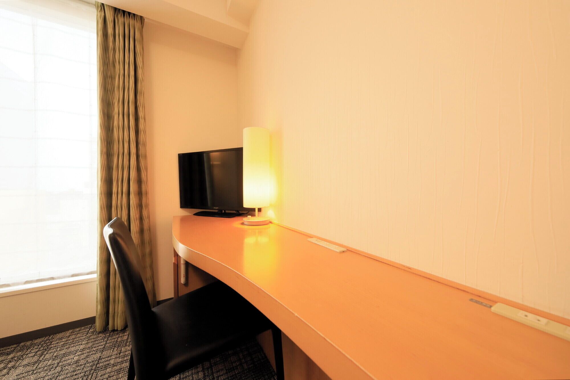 Quarto de hotel com escrivaninha de madeira, televisão, luminária e uma cadeira.