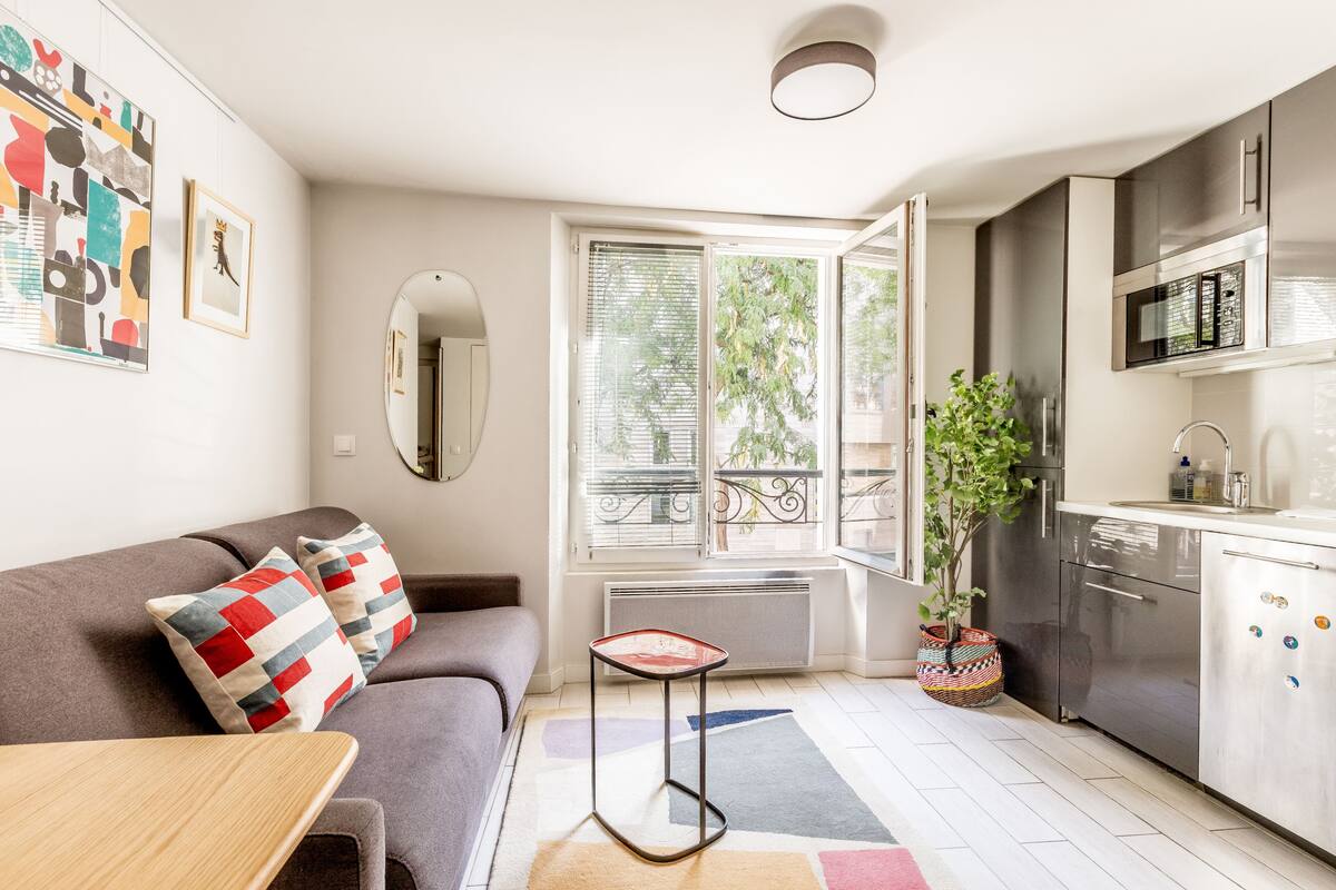Um espaço compacto com sofá, mesa pequena, cozinha equipada com frigorífico e micro-ondas, e janela com vista para o verde.