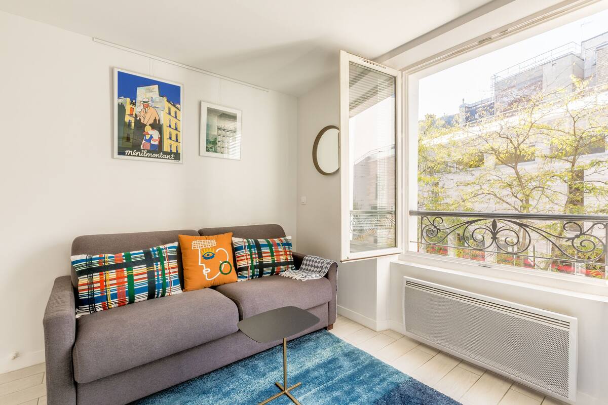 Sala de estar com um sofá cinza, almofadas coloridas, uma mesa pequena e uma janela com vista para edifícios e árvores.