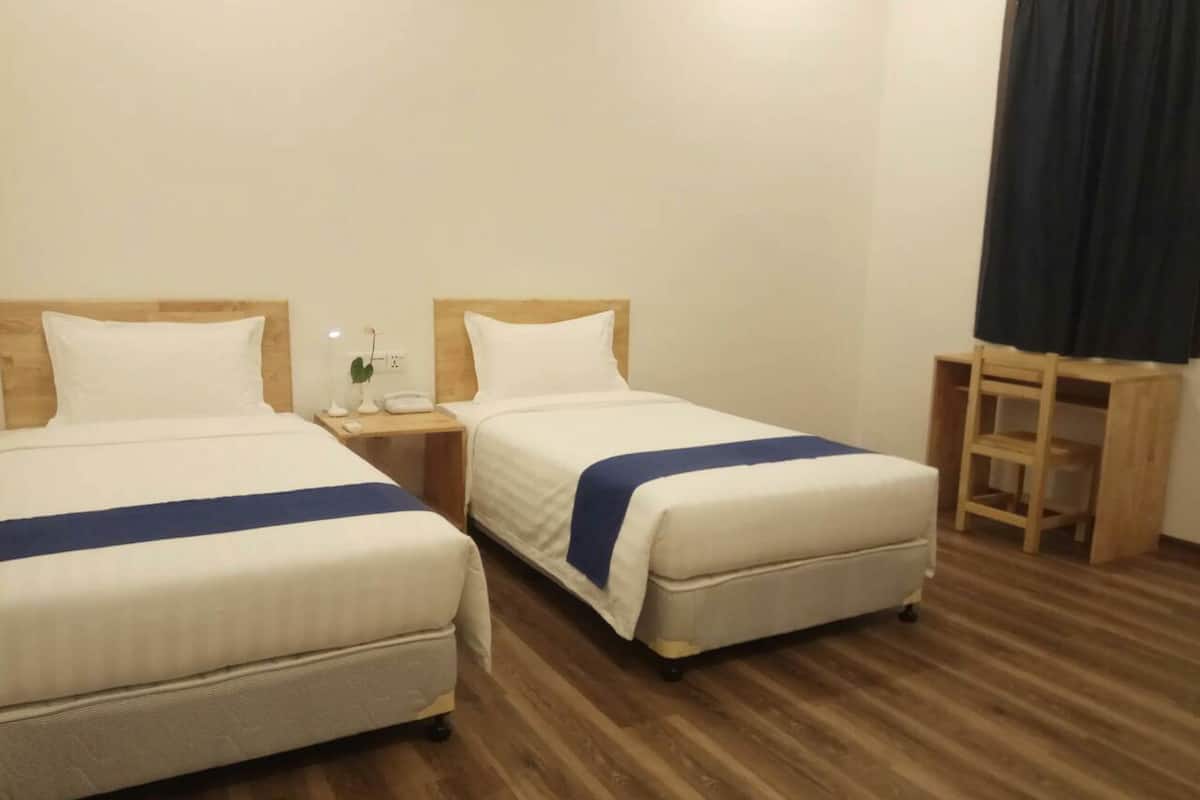 Quarto de hotel com duas camas, cabeceiras de madeira, mesa de cabeceira com abajur e uma pequena mesa de madeira.