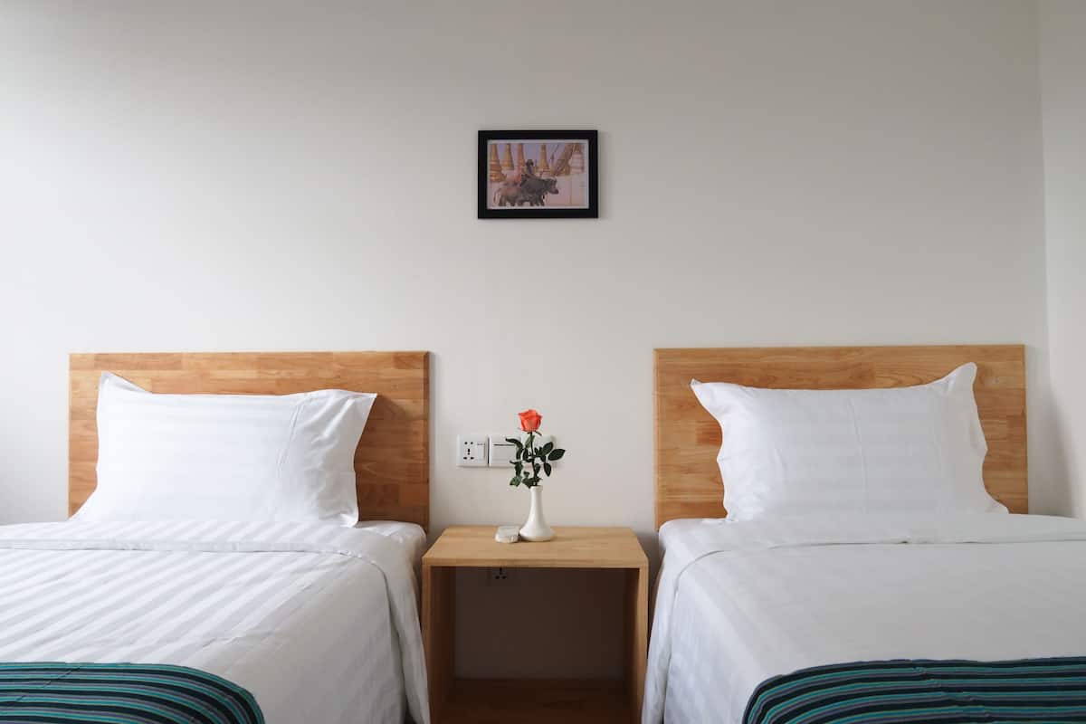 Quarto de hotel com duas camas, uma mesa de cabeceira com um vaso com uma única rosa e um quadro na parede.