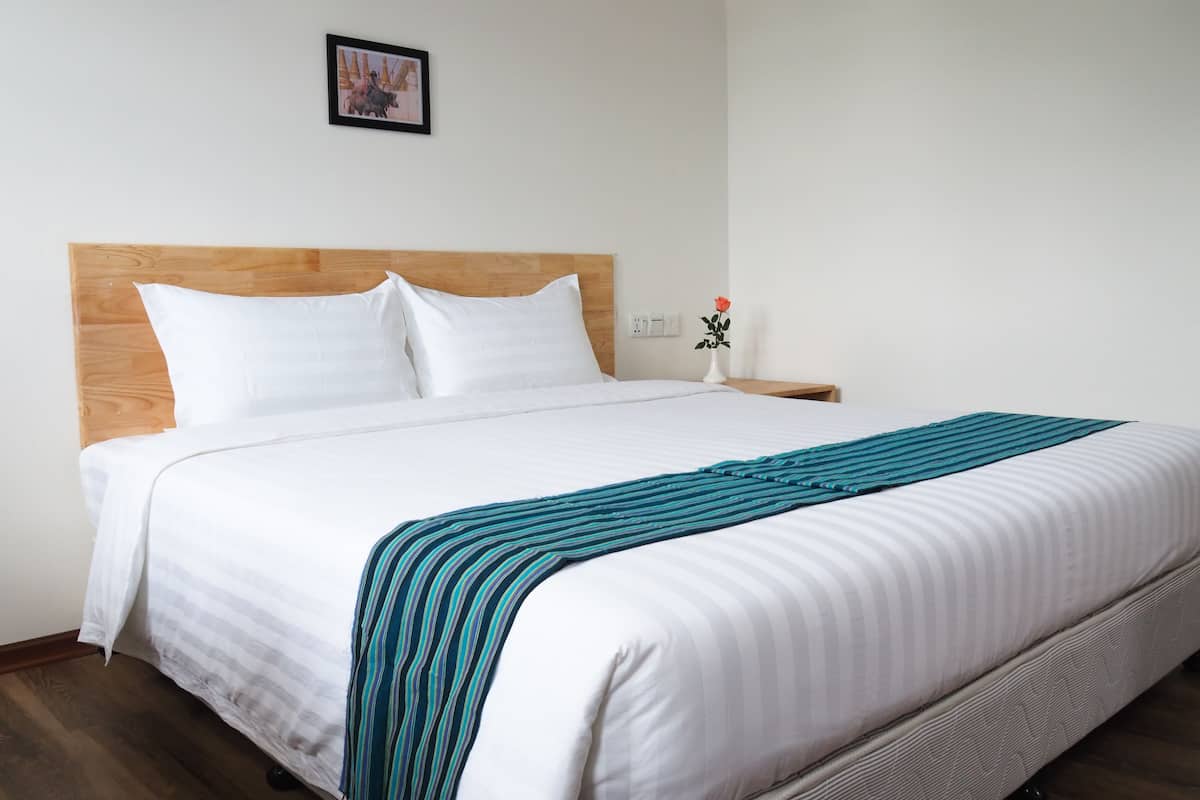 Cama bem arrumada com roupa de cama branca e manta listrada azul-turquesa, cabeceira de madeira, mesa de cabeceira pequena com um vaso de flores e um quadro na parede.