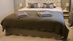 Quarto com cabeceira estofada, cama grande com colcha escura, duas almofadas e mesa de cabeceira com abajur.