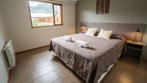 Quarto com uma cama grande, janela com vista para edifícios e montanhas, radiador e mesa de cabeceira com abajur.