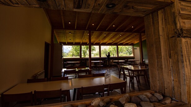 Uma sala de jantar rústica com mesas e bancos de madeira, janelas amplas e vista para o verde lá fora.