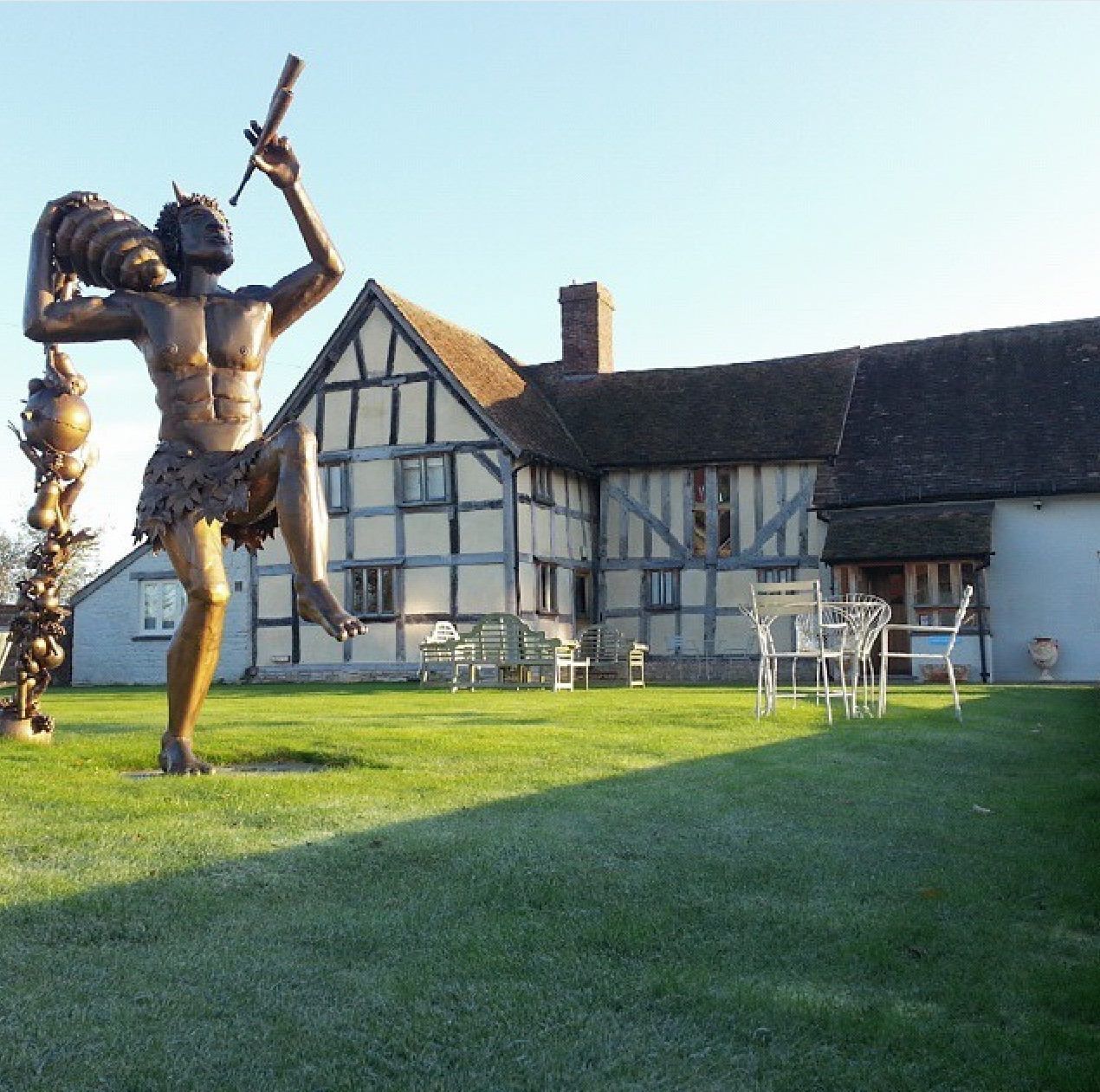 Estátua de uma figura musculosa segurando uma lança em frente a um edifício em estilo Tudor, com gramado e área externa para sentar.
