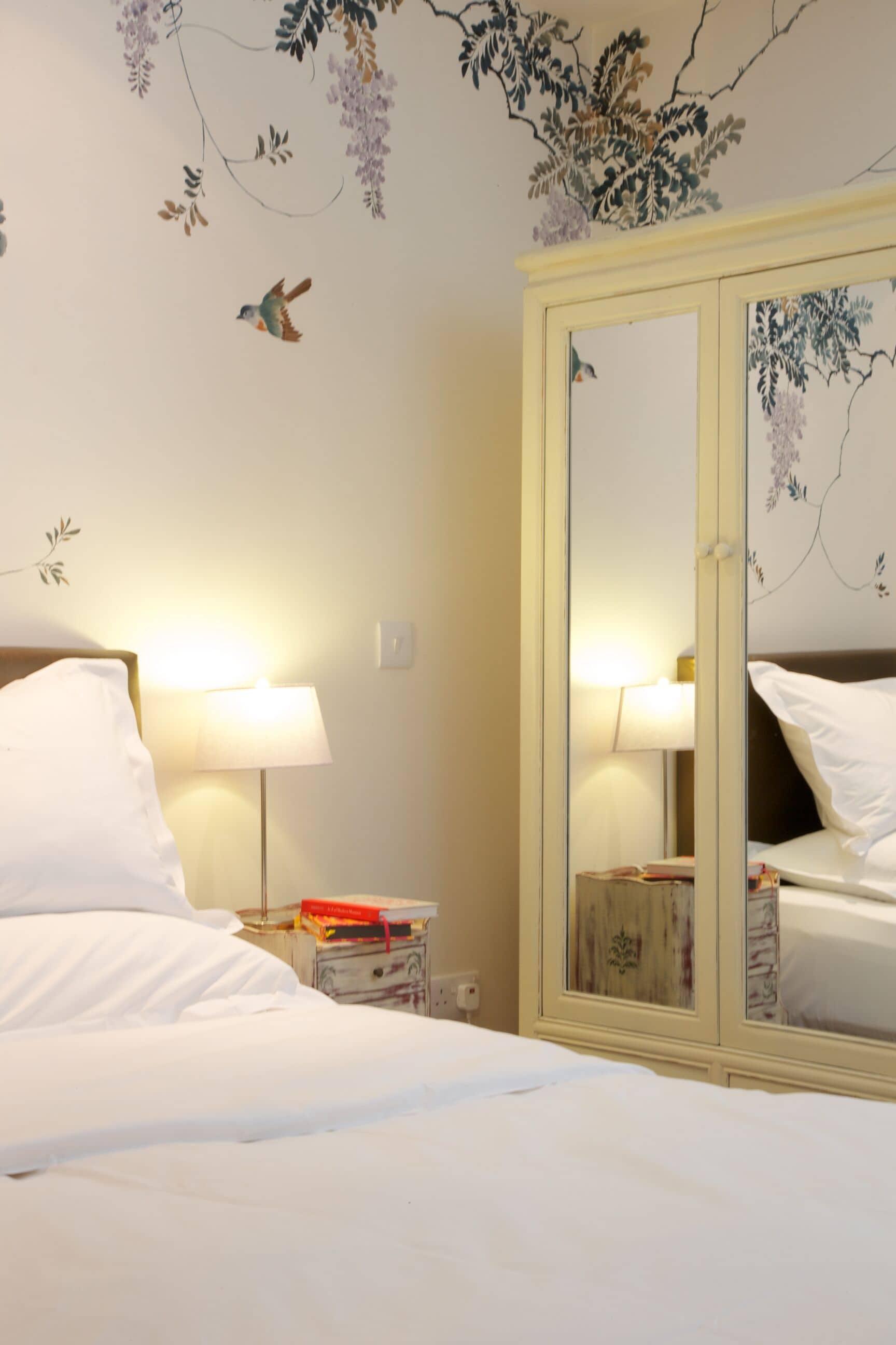 Quarto de hotel com cama, duas luminárias de cabeceira, mesa de cabeceira com livros, espelho e decorações nas paredes.