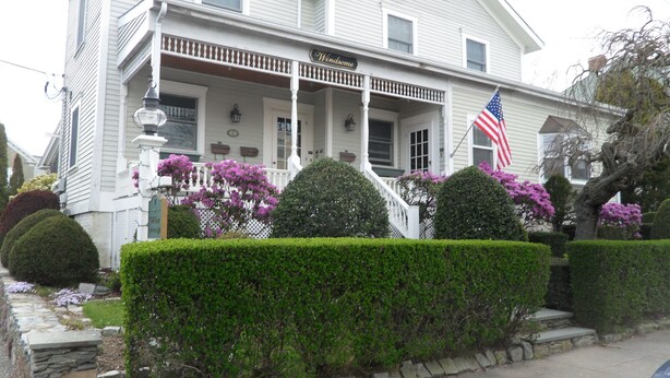 Uma casa de dois andares com varanda, uma bandeira americana e sebes bem cuidadas.