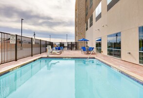 Piscina com espreguiçadeiras e guarda-sóis, cercada por uma vedação e um edifício com grandes janelas.