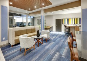 Um hall de hotel moderno com carpete azul, sofá bege e uma pequena mesa com cadeiras.