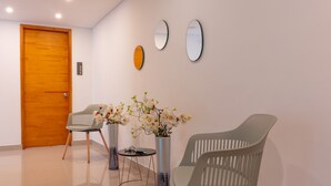 Uma área de recepção com porta de madeira, duas cadeiras cinzas, uma pequena mesa com um vaso de flores e três espelhos fixados na parede.