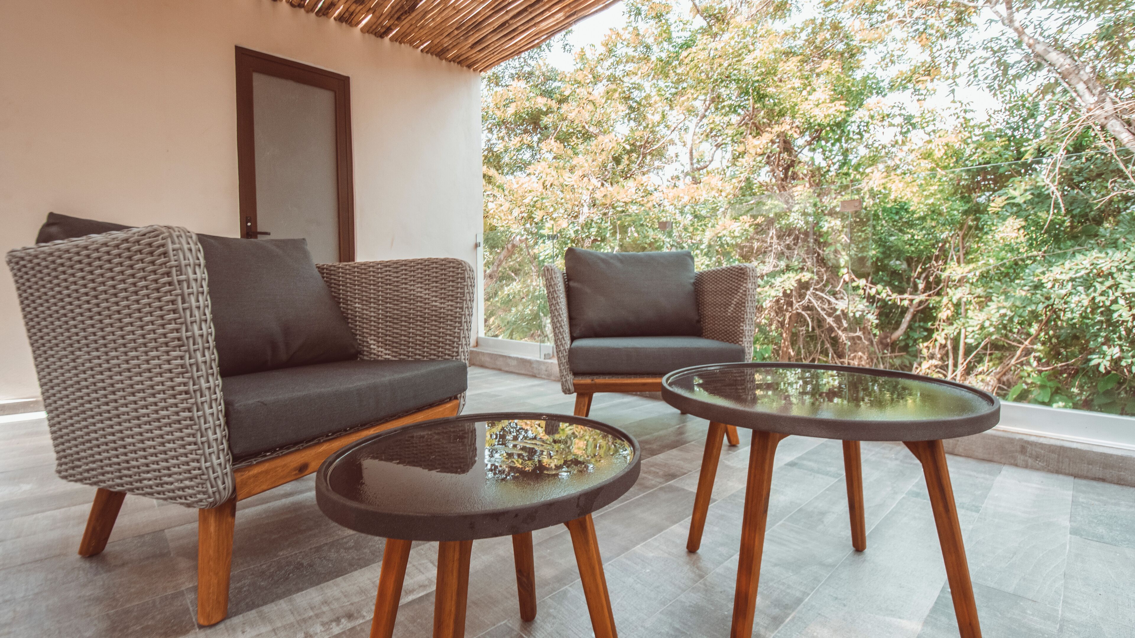 Uma área de estar moderna ao ar livre, com um sofá e duas poltronas, mesa de vidro e vista para árvores.