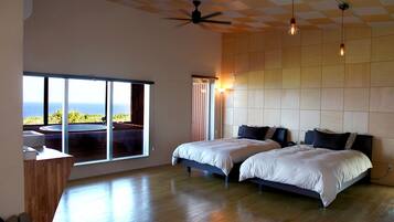 Um quarto espaçoso com duas camas, uma janela ampla com vista para o mar, um ventilador de teto e um abajur.