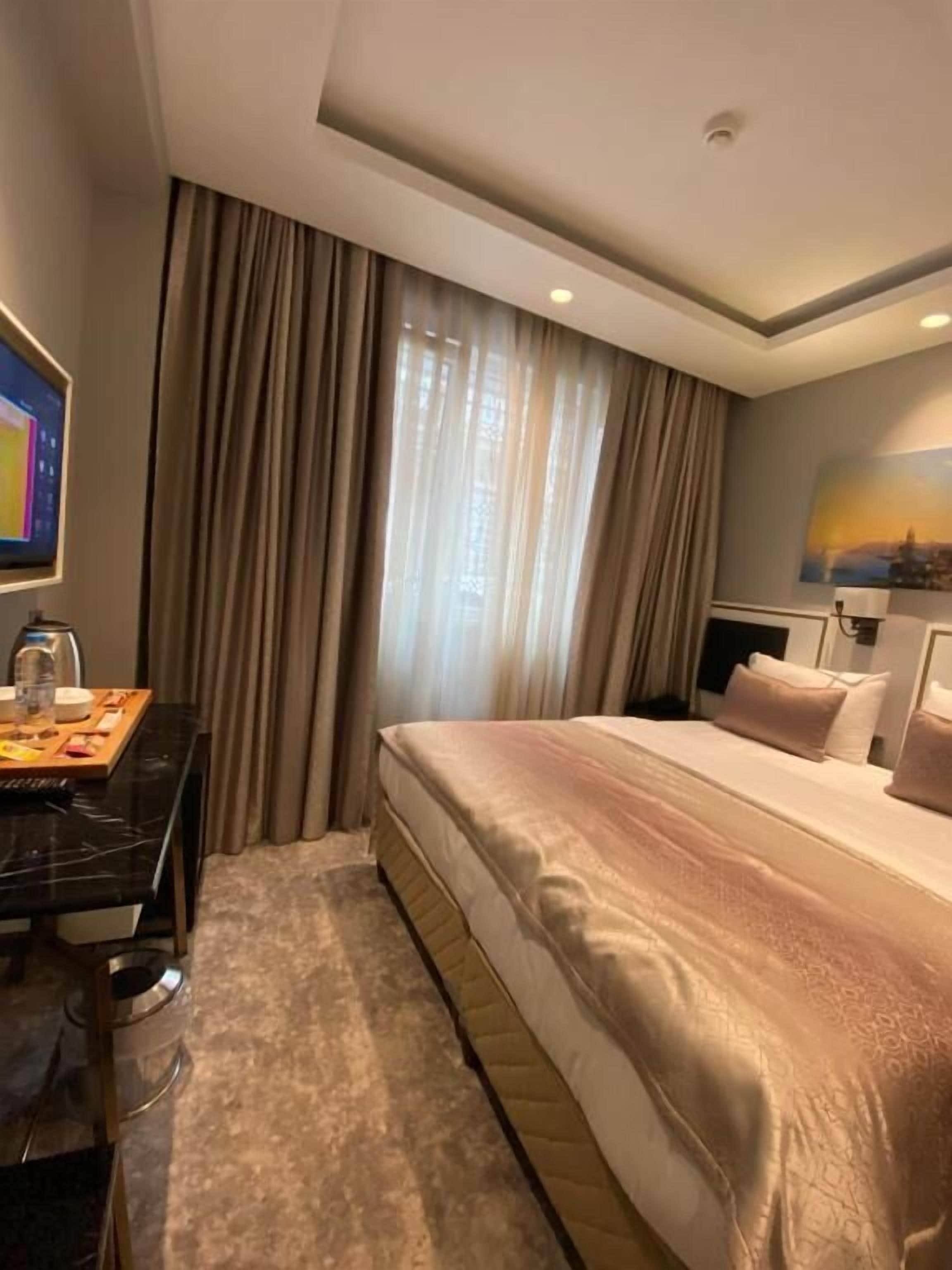 Quarto de hotel com uma cama grande, uma escrivaninha com computador e uma janela com cortinas.