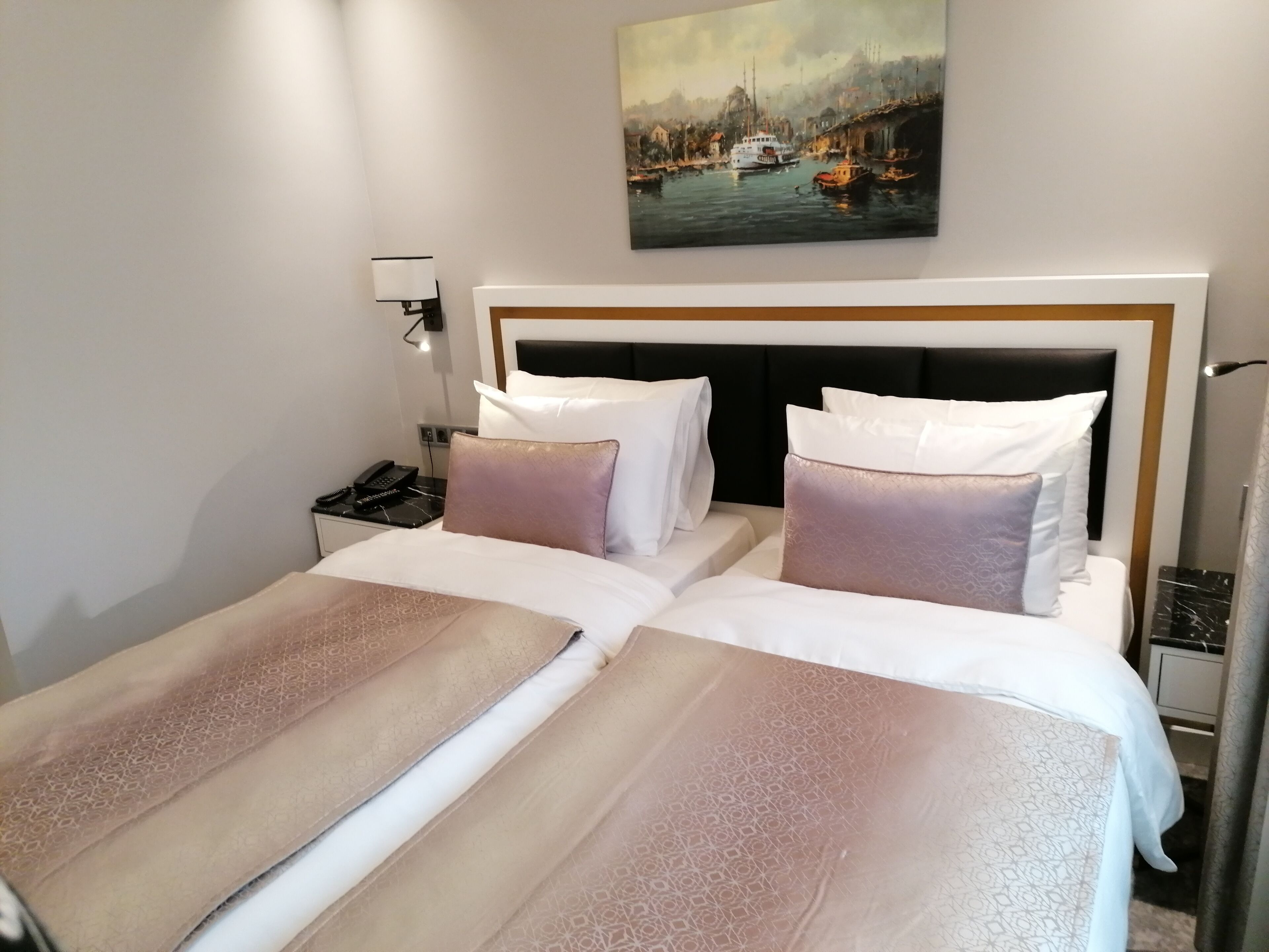Quarto de hotel com uma cama grande, uma mesa de cabeceira, um quadro de uma paisagem portuária e uma luminária.