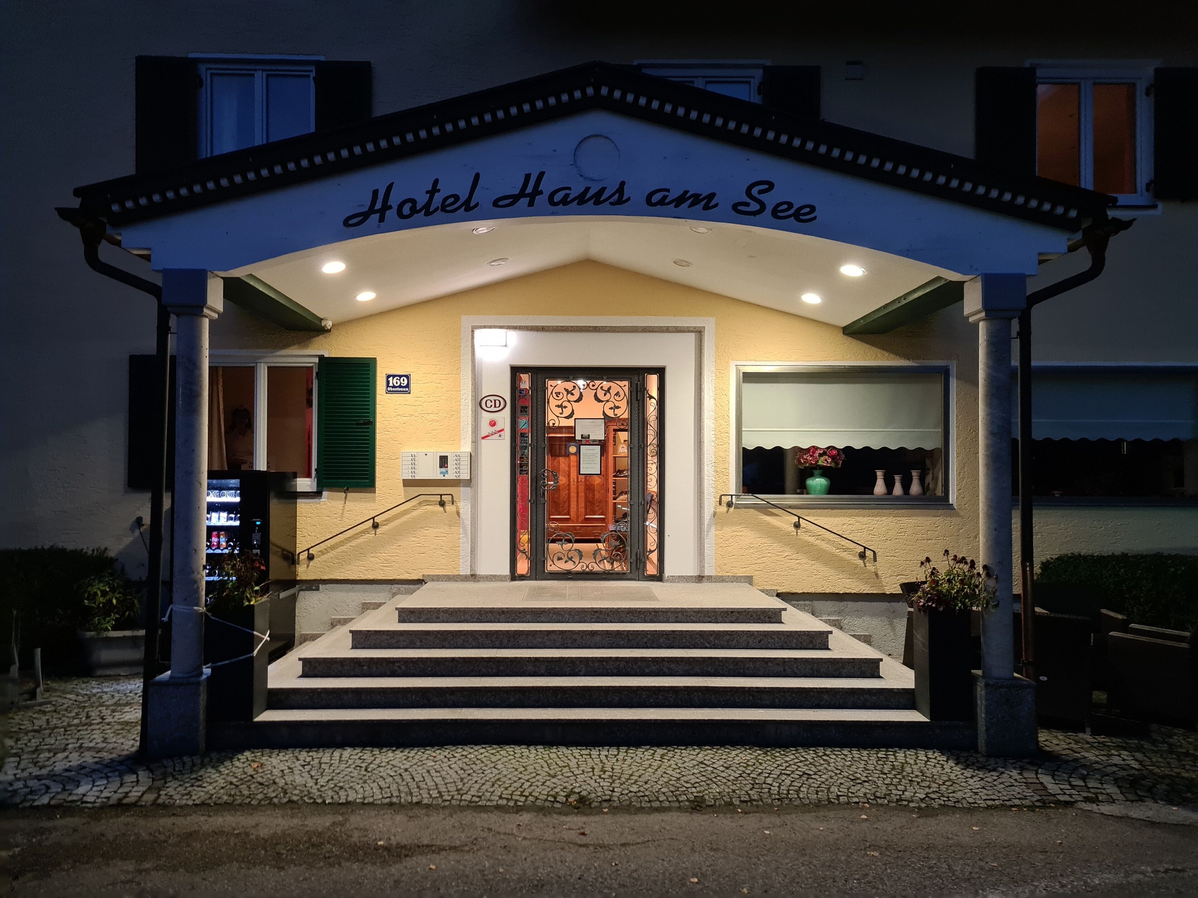 Entrada de um hotel com uma placa indicando "Hotel Haus am See", degraus que levam à porta e uma pequena área externa com um vaso de planta.