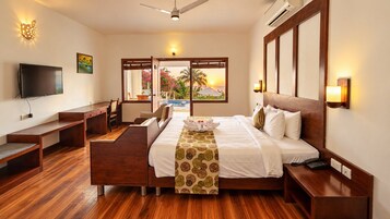 Quarto de hotel com cama, mesas de cabeceira, escrivaninha, poltrona, televisão e janela com vista para uma piscina e palmeiras.
