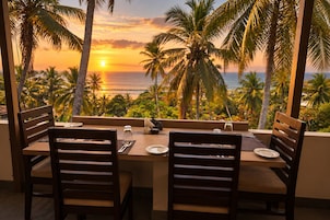 Mesa posta para dois com vista para o pôr do sol sobre o oceano, rodeada por palmeiras.