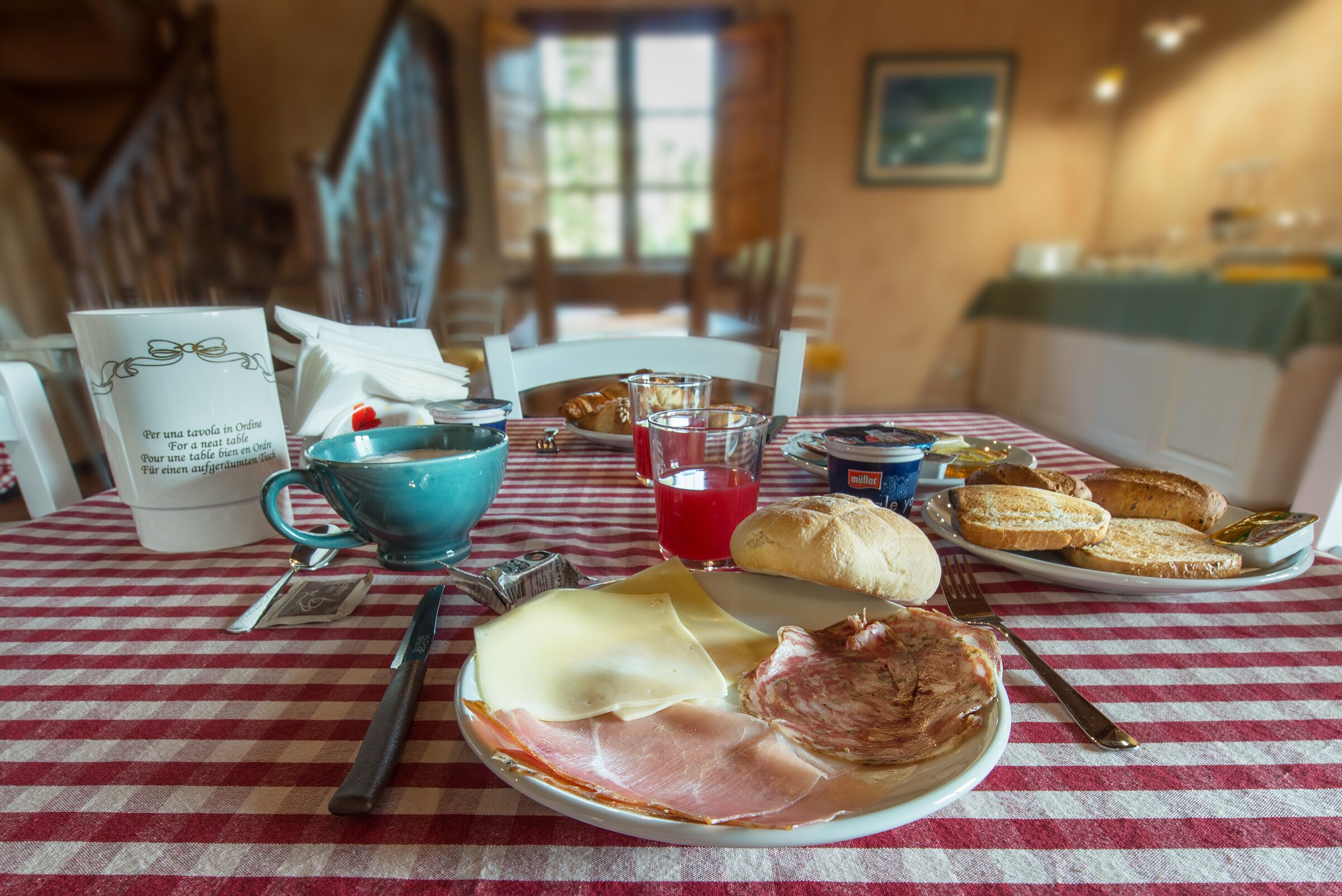 Mesa de café da manhã com um prato de presunto, queijo e pão, uma xícara azul e um copo de suco vermelho.