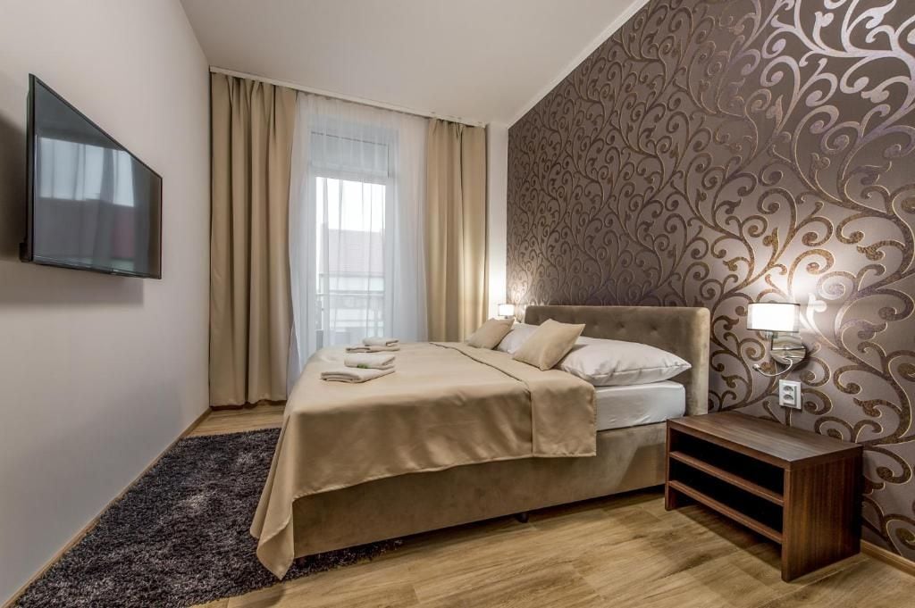 Quarto com cama, televisão fixada na parede, mesa de cabeceira com abajur e parede estampada.