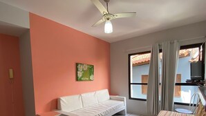 Um quarto pequeno e bem iluminado, com um sofá branco, uma cama com colcha listrada, uma mesa de jantar com toalha de mesa florida e um ventilador de teto.
