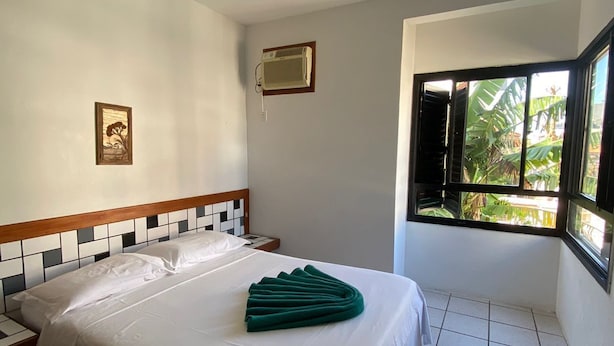 Quarto de hotel com cama, mesa de cabeceira, ar condicionado fixado na parede, quadro emoldurado e janela com persiana aberta.