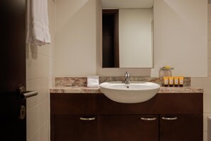 Um banheiro com pia branca, bancada de mármore e espelho acima. Há produtos de higiene no balcão e uma toalha pendurada na parede.