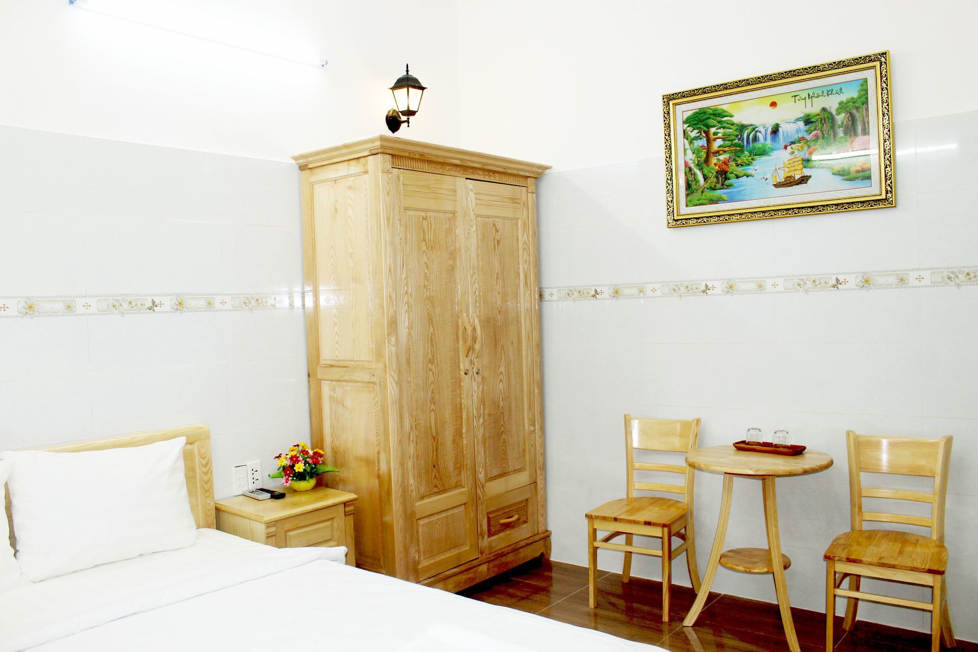 Quarto de hotel com cama, guarda-roupa de madeira, mesa de cabeceira com abajur, pequena mesa com duas cadeiras e quadro na parede.