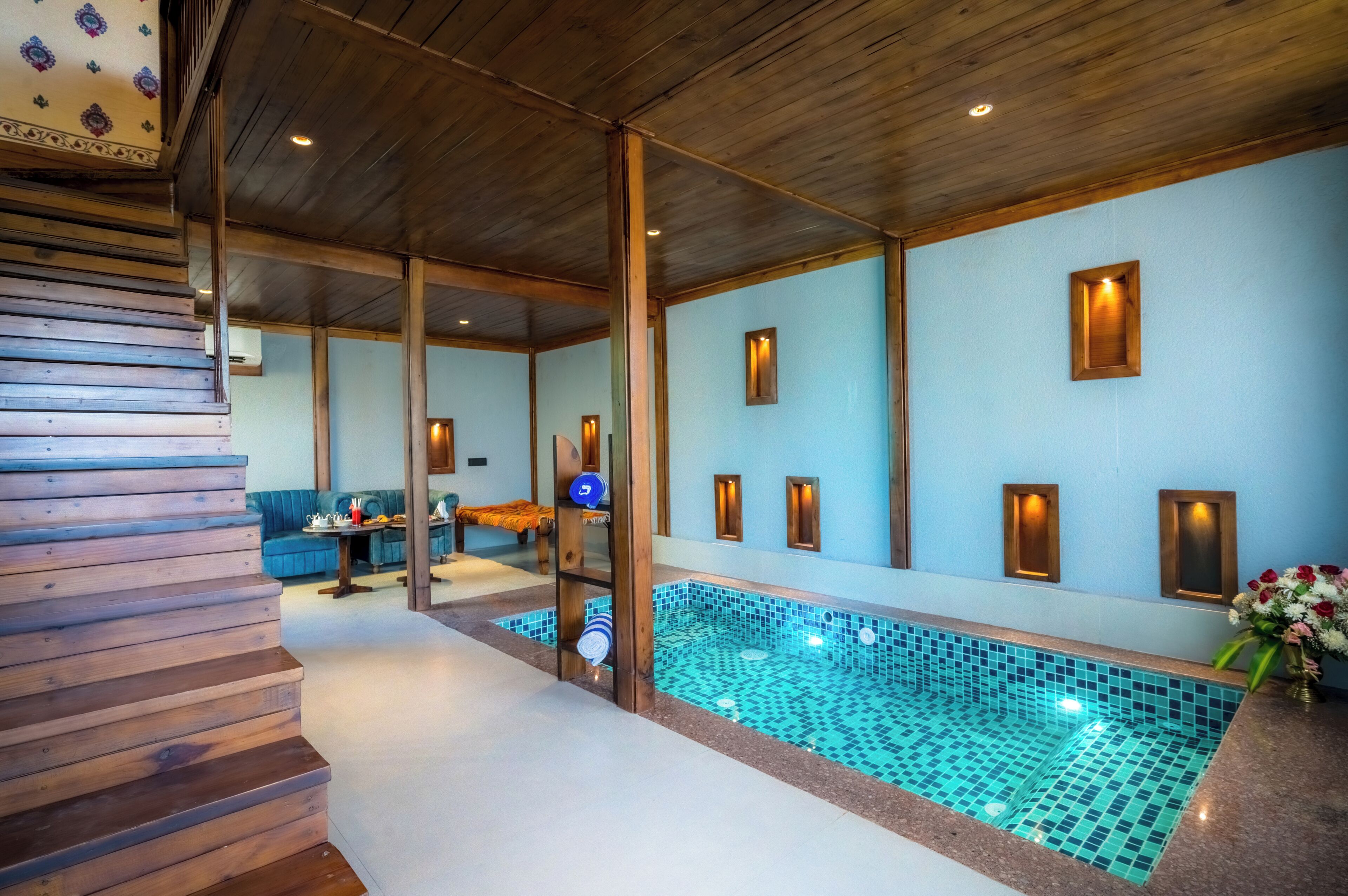 Uma área de spa com escada de madeira, mesa de massagem, pequena piscina e parede azul com iluminação embutida.