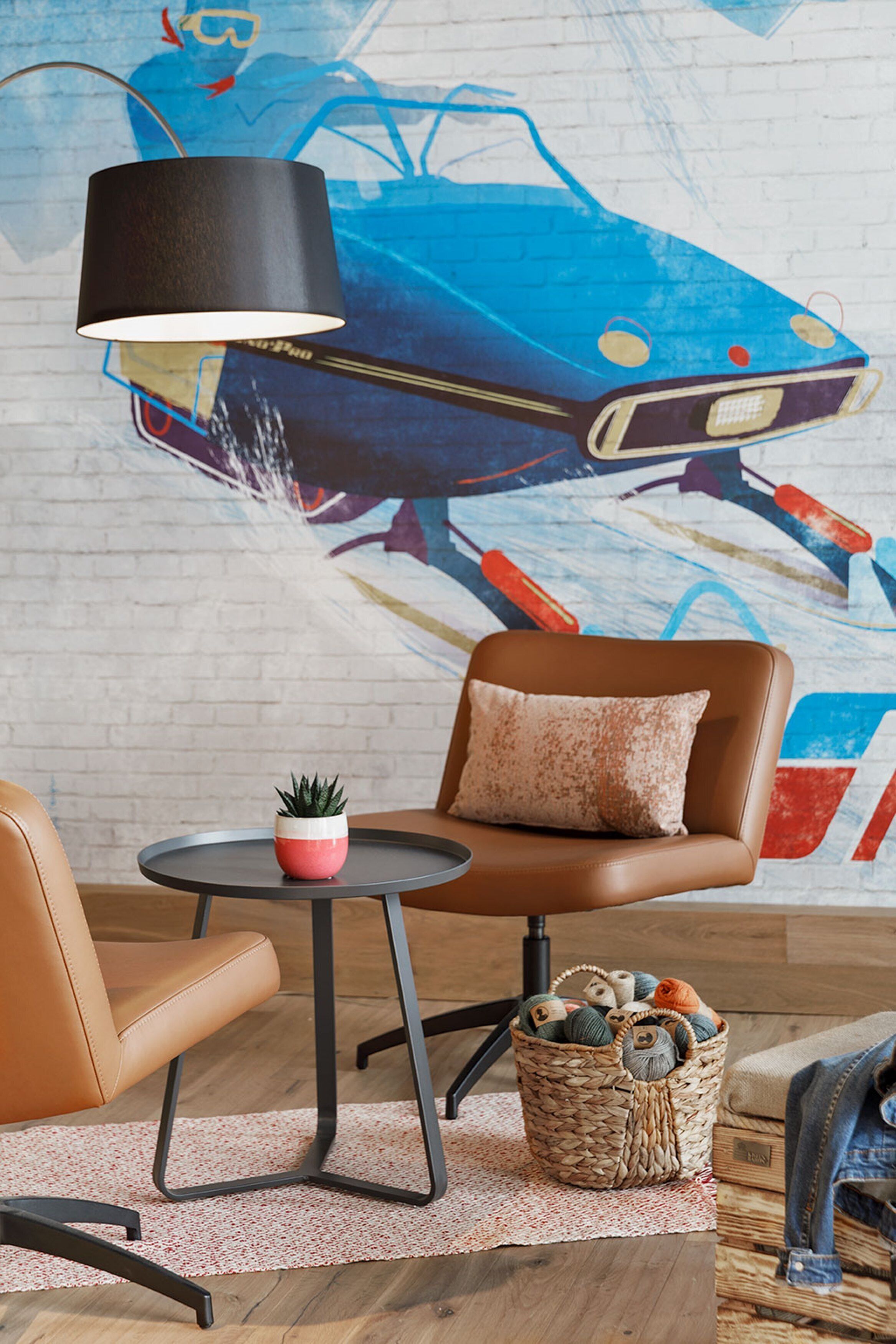 Um quarto com uma poltrona de couro marrom, uma pequena mesa redonda com abajur, uma cesta de palha com lã e um mural de um carro azul na parede.
