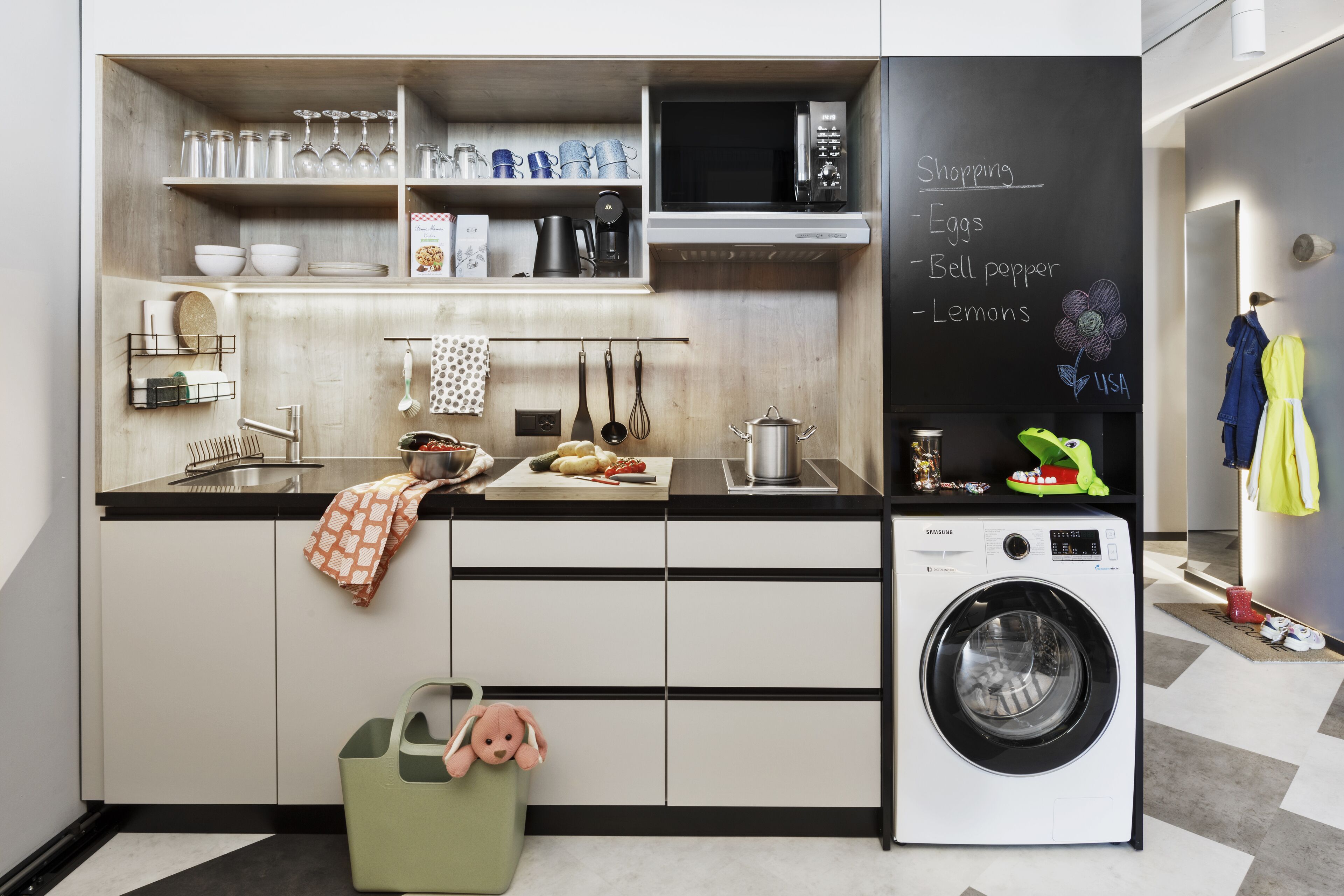 Cozinha moderna com máquina de lavar e secar integradas, eletrodomésticos de aço inoxidável e quadro negro com lista de compras.