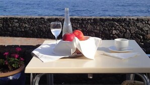 Uma mesa com uma garrafa, um copo e uma cesta de frutas, junto a uma parede de pedra com vista para o mar.