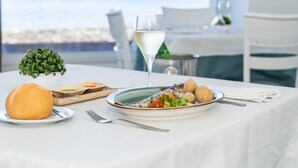 Uma mesa com refeição, um copo de vinho branco e vista para o mar.