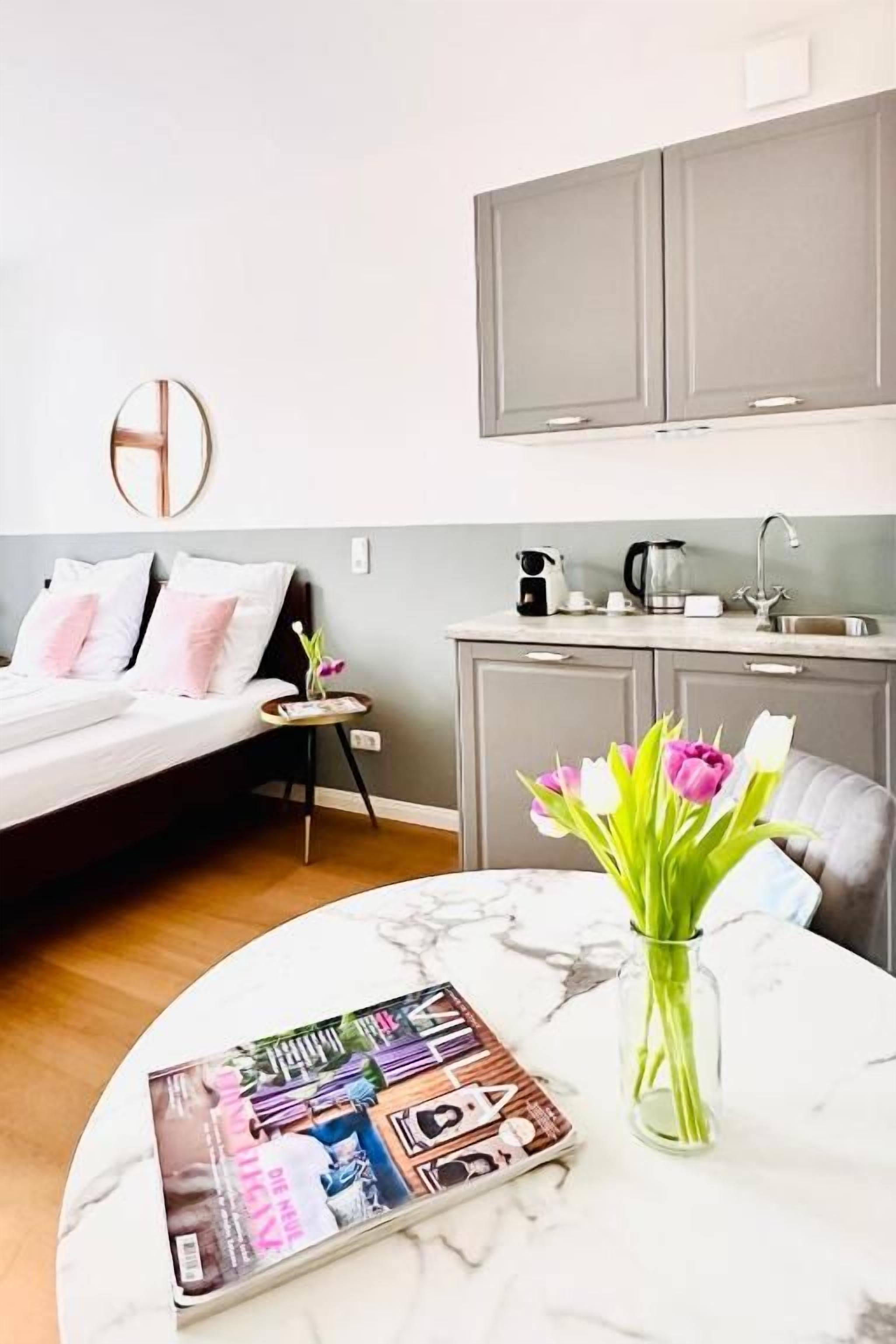 Quarto moderno com cama, uma mesinha com revista e vaso, cozinha compacta com armários e parede cinza.