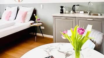 Quarto moderno com cama, uma mesinha com revista e vaso, cozinha compacta com armários e parede cinza.