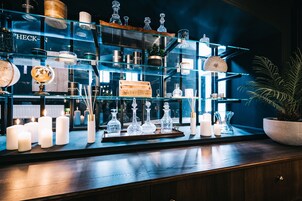 Uma área de bar com prateleiras exibindo vários frascos e copos, uma planta em vaso e velas.
