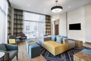 Quarto de hotel moderno com sofá modular, poltronas, televisão de tela plana e janelas amplas com cortinas.
