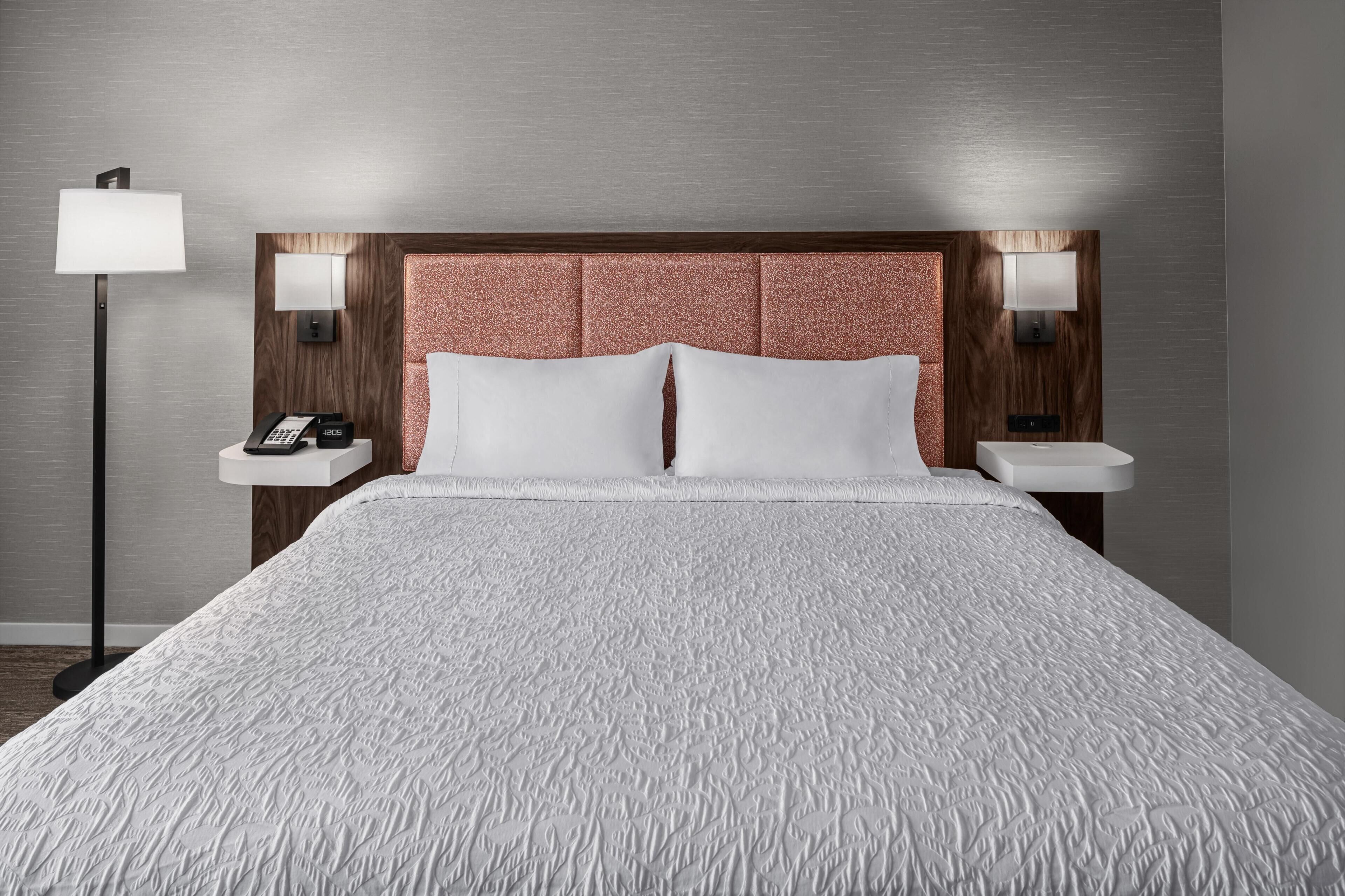 Quarto de hotel com uma cama grande, duas mesas de cabeceira, um criado-mudo, uma lâmpada e um telefone fixado na parede.