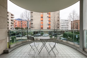 Varanda com mesa e cadeiras, com vista para um parque de estacionamento e edifícios.