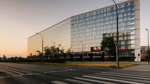 Um edifício moderno de vidro com fachada plana, situado no cruzamento de uma rua com faixas de pedestres e semáforos.