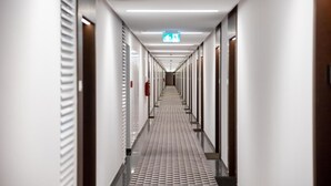 Um corredor comprido e estreito com piso estampado, portas em ambos os lados e um sinal de saída azul no teto.