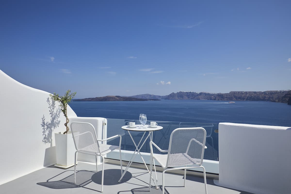 Varanda com duas cadeiras de metal branco e uma mesa redonda pequena, com vista para o mar azul límpido e montanhas ao longe.