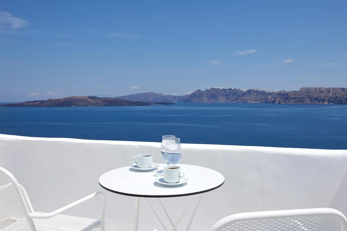 Uma mesa branca com duas xícaras de café e um copo d'água, com vista para um mar azul cristalino e falésias ao longe.