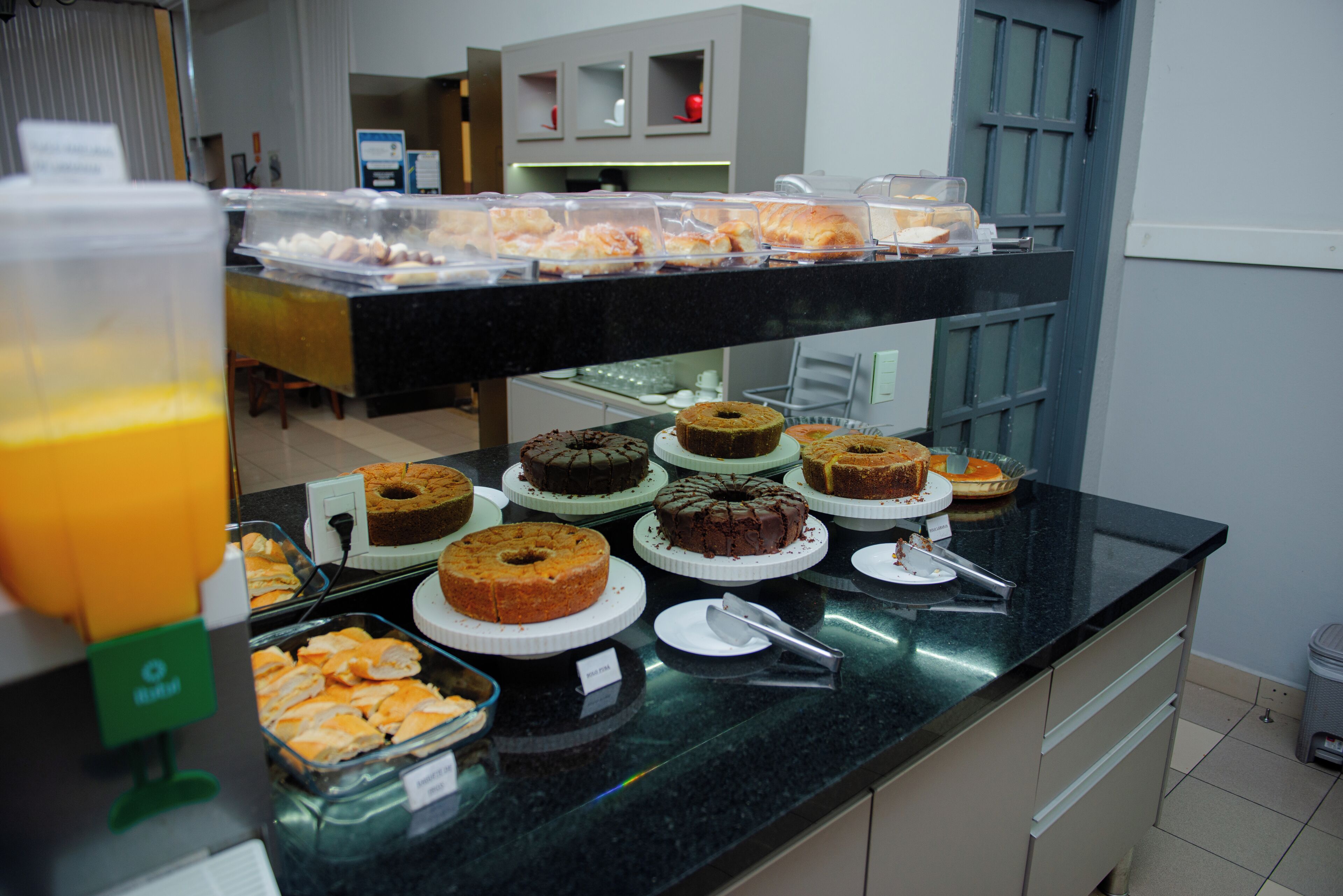 Buffet com diversos bolos e doces, um dispensador de suco e um aviso indicando o preço.