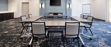 Uma sala de conferências com uma mesa grande, cadeiras, uma TV e um quadro branco.