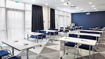 Sala de conferências com mesas, cadeiras, um projetor e uma janela grande com cortinas.