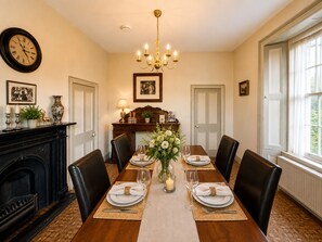 Uma sala de jantar com mesa de madeira posta para quatro pessoas, um lustre e uma lareira.