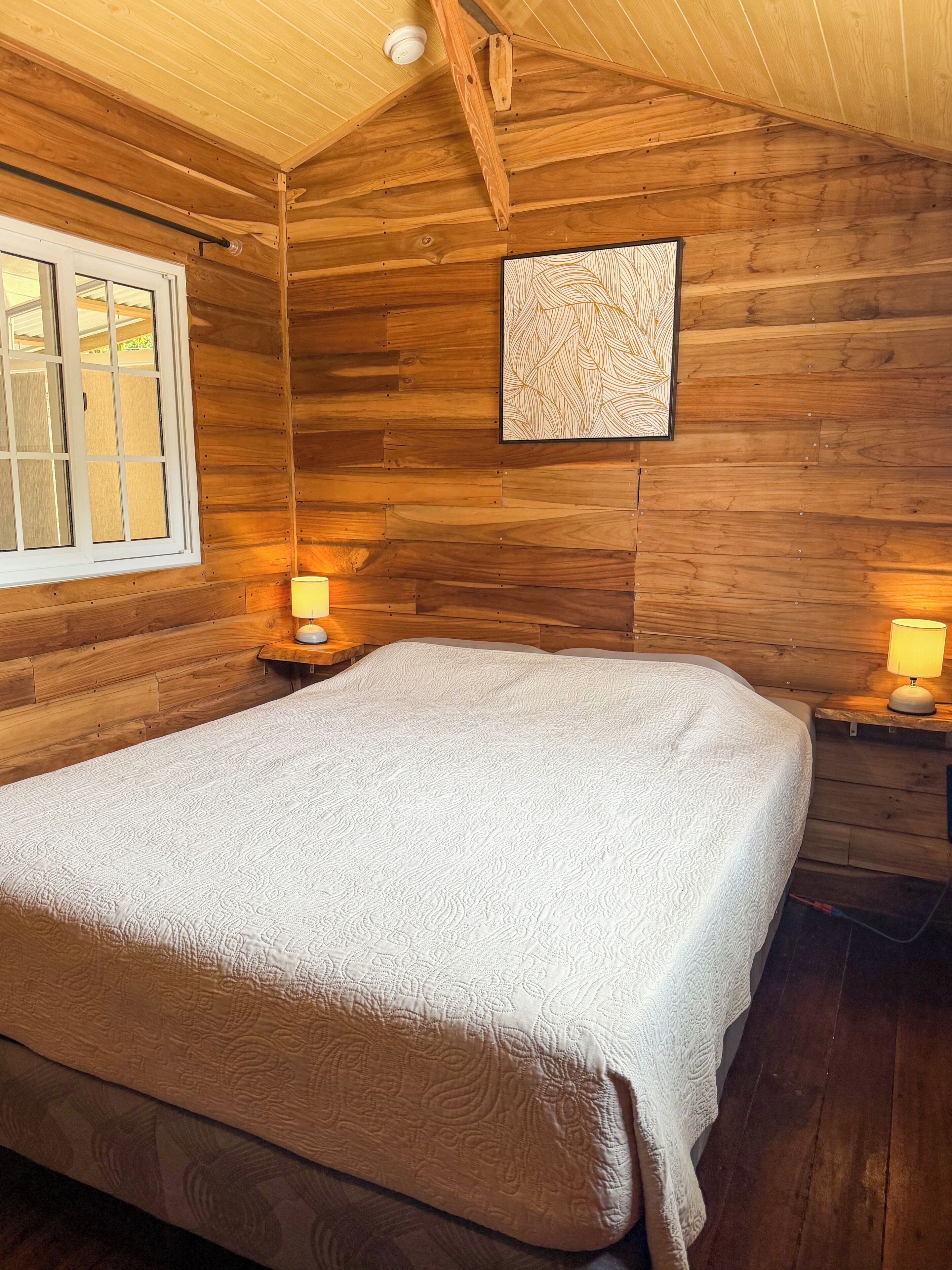 Quarto de cabana de madeira com uma cama, duas luminárias de cabeceira, uma janela e um quadro na parede.