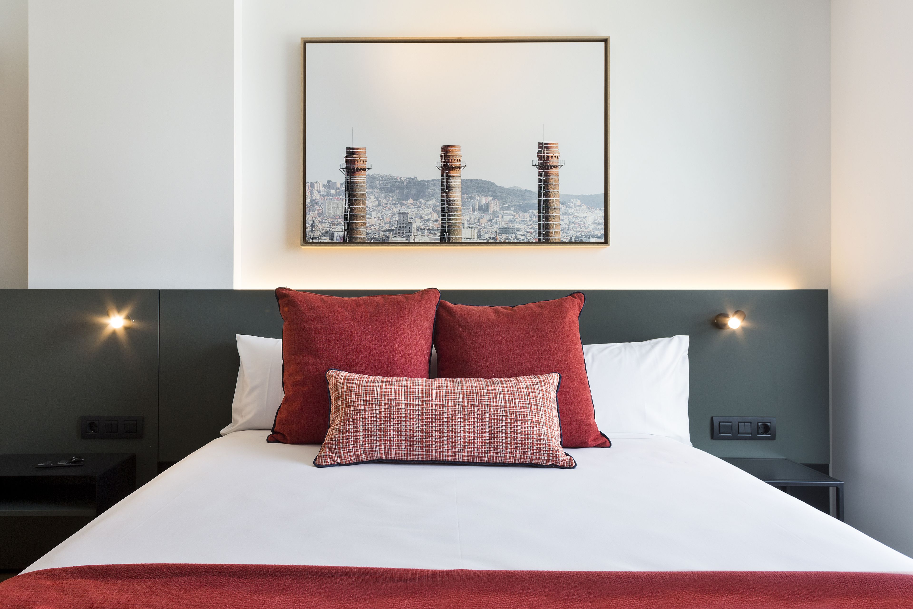 Quarto de hotel com cama, almofadas vermelhas e brancas, vista para a cidade e uma moldura na parede.